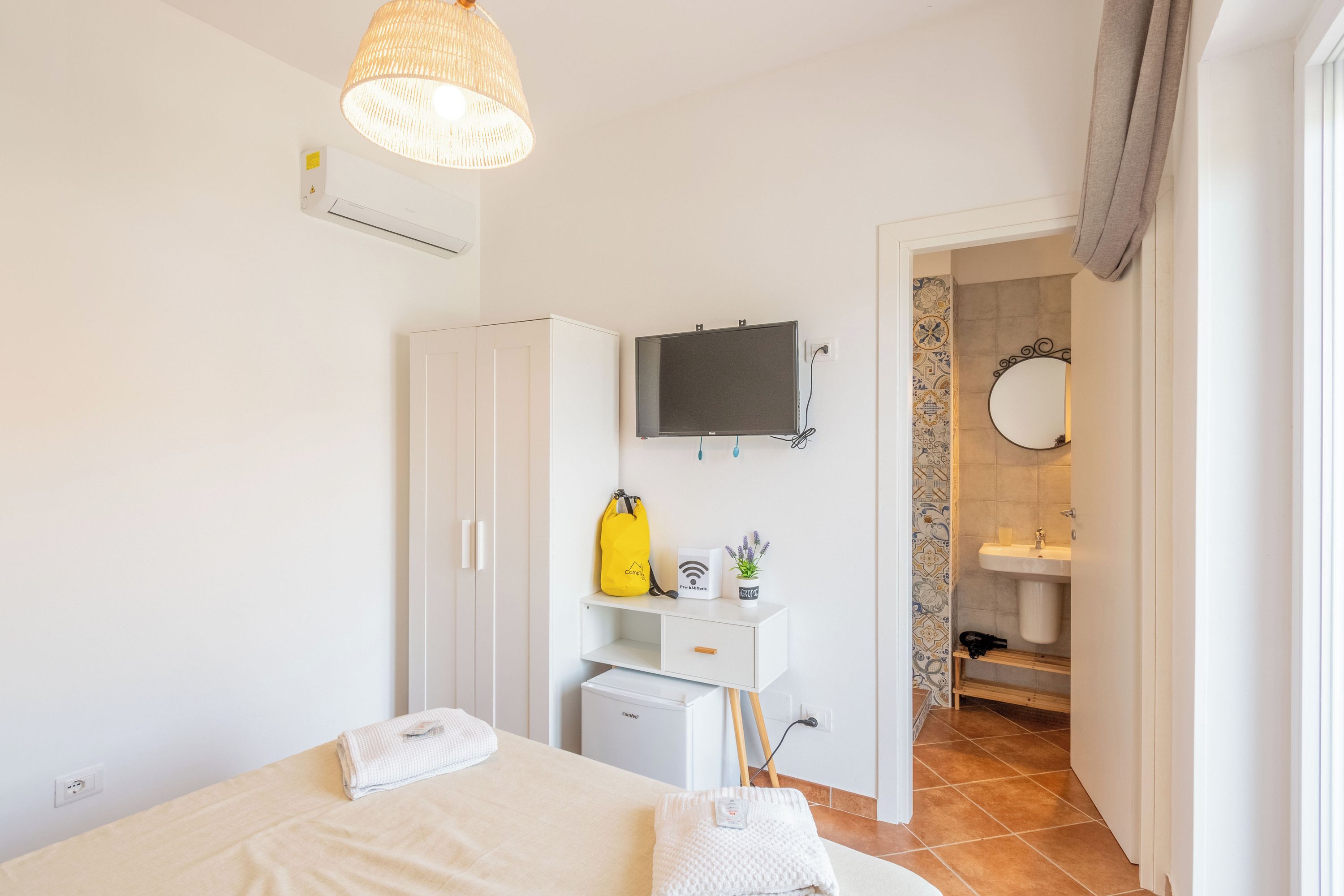 Quarto de hotel com cama, televisão, guarda-roupa, pequena escrivaninha e banheiro visível através de uma porta aberta.