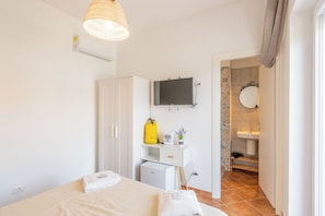 Quarto de hotel com cama, televisão, guarda-roupa, pequena escrivaninha e banheiro visível através de uma porta aberta.