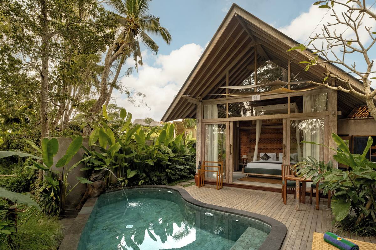 Uma villa de madeira com piscina, um quarto com cama e cortinas, e vegetação tropical exuberante.