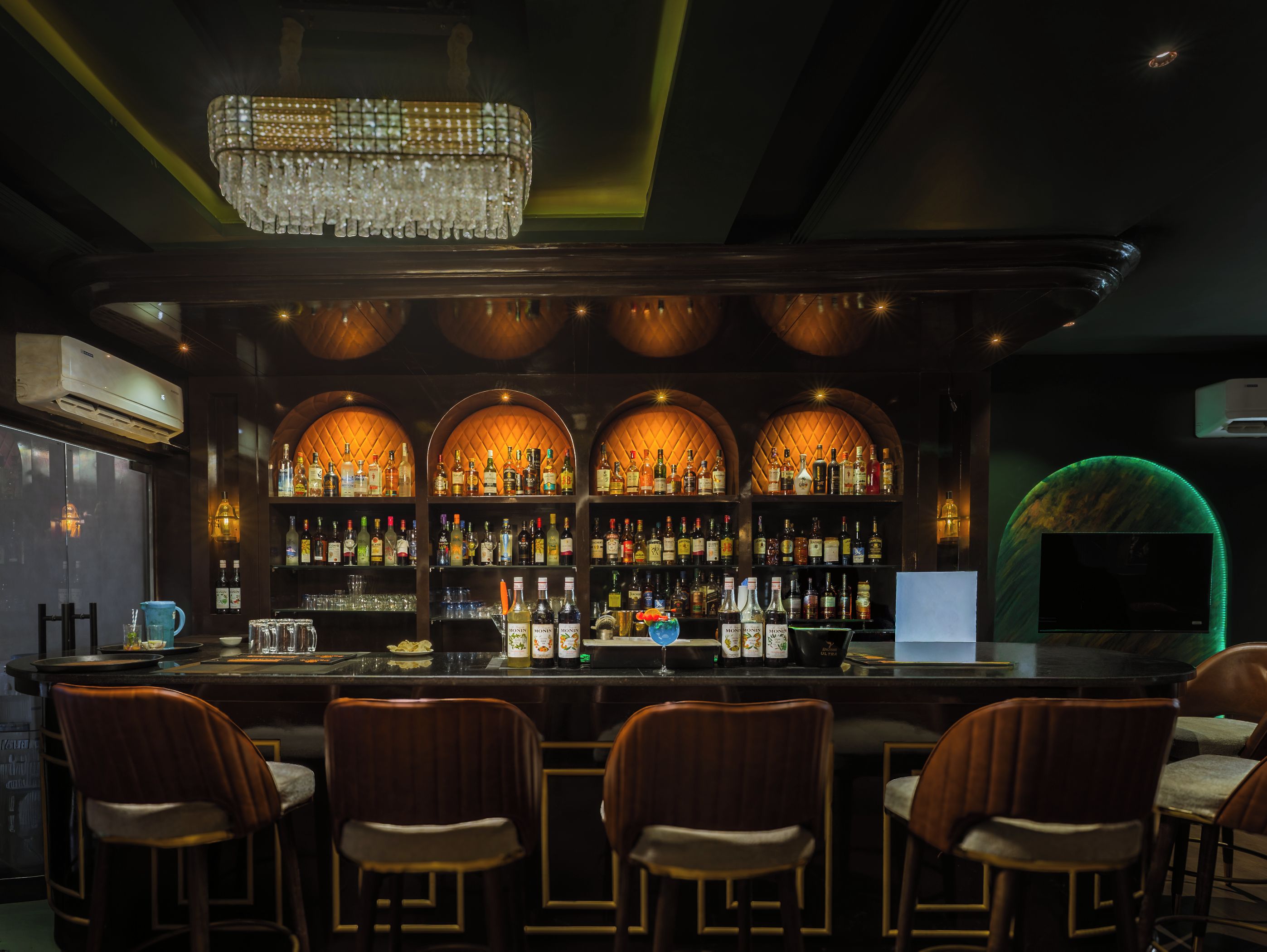 Uma área de bar com um candelabro, estantes arqueadas com garrafas de bebidas e banquetas.