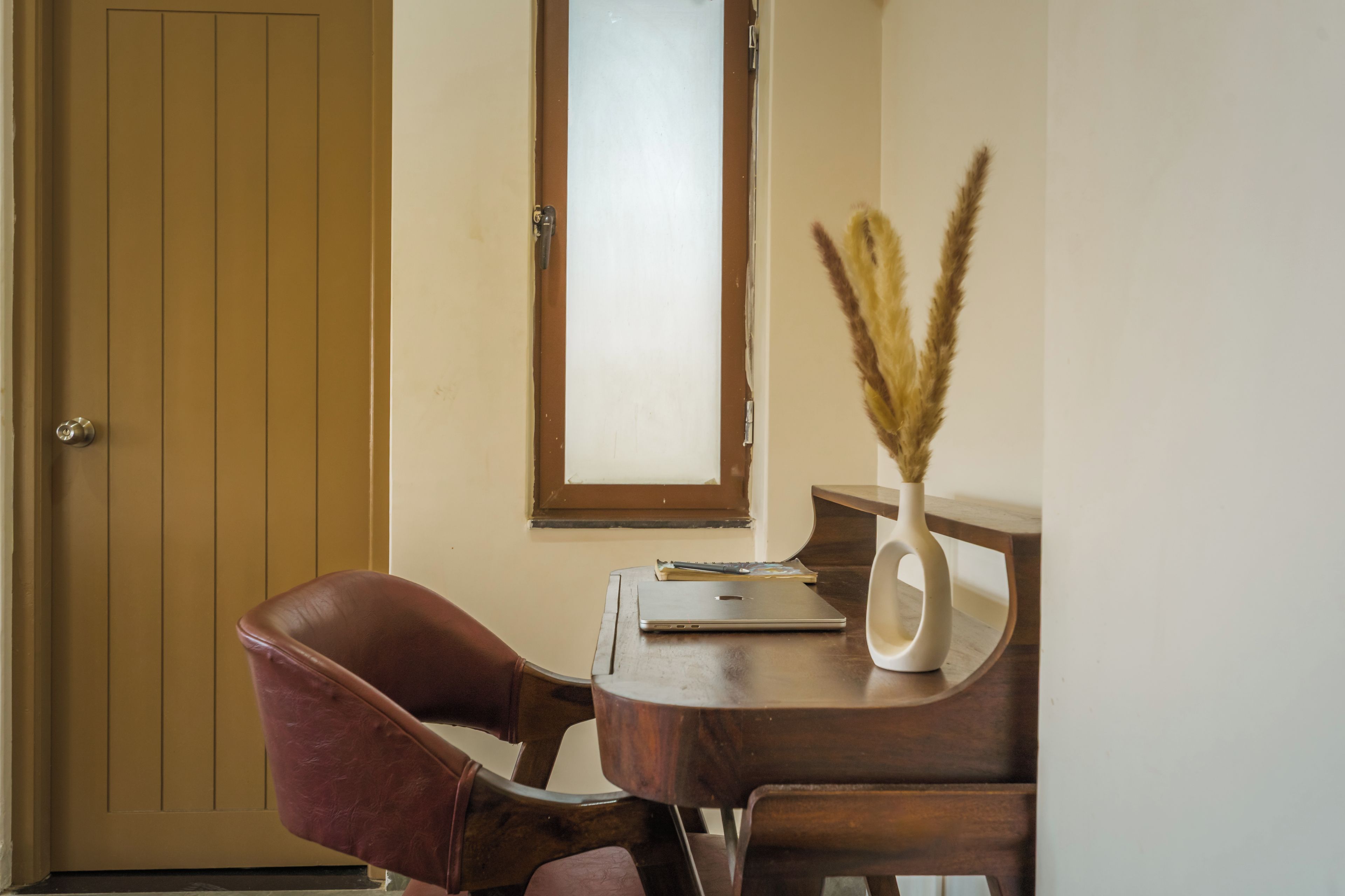 Quarto com porta de madeira, uma escrivaninha com um vaso de capim-do-campo e uma cadeira.