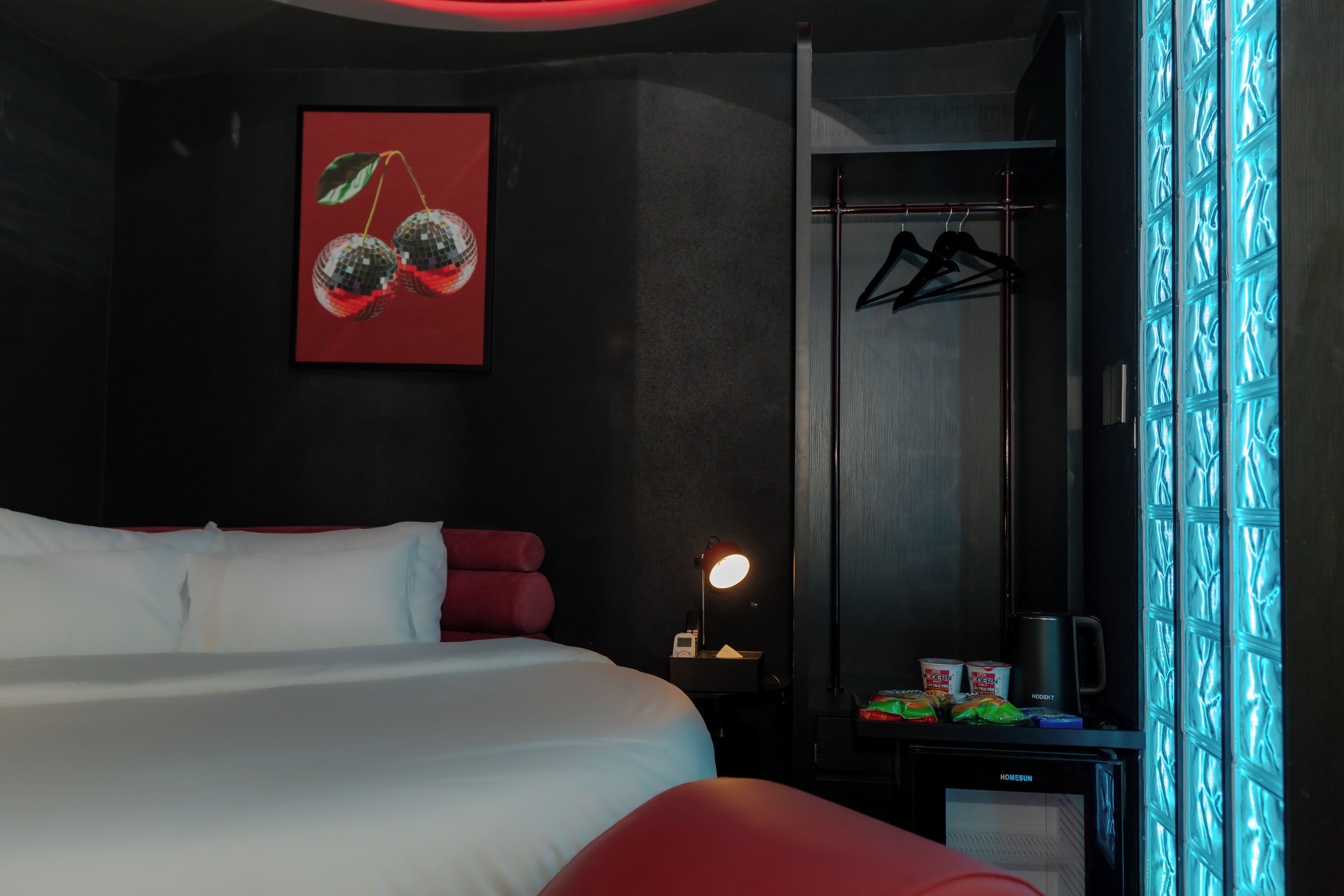 Quarto de hotel com uma cama, uma escrivaninha com abajur, um cabideiro e uma janela com cortinas estampadas.