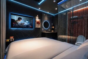 Quarto de hotel moderno com uma cama grande, televisão fixada na parede e luminária circular.