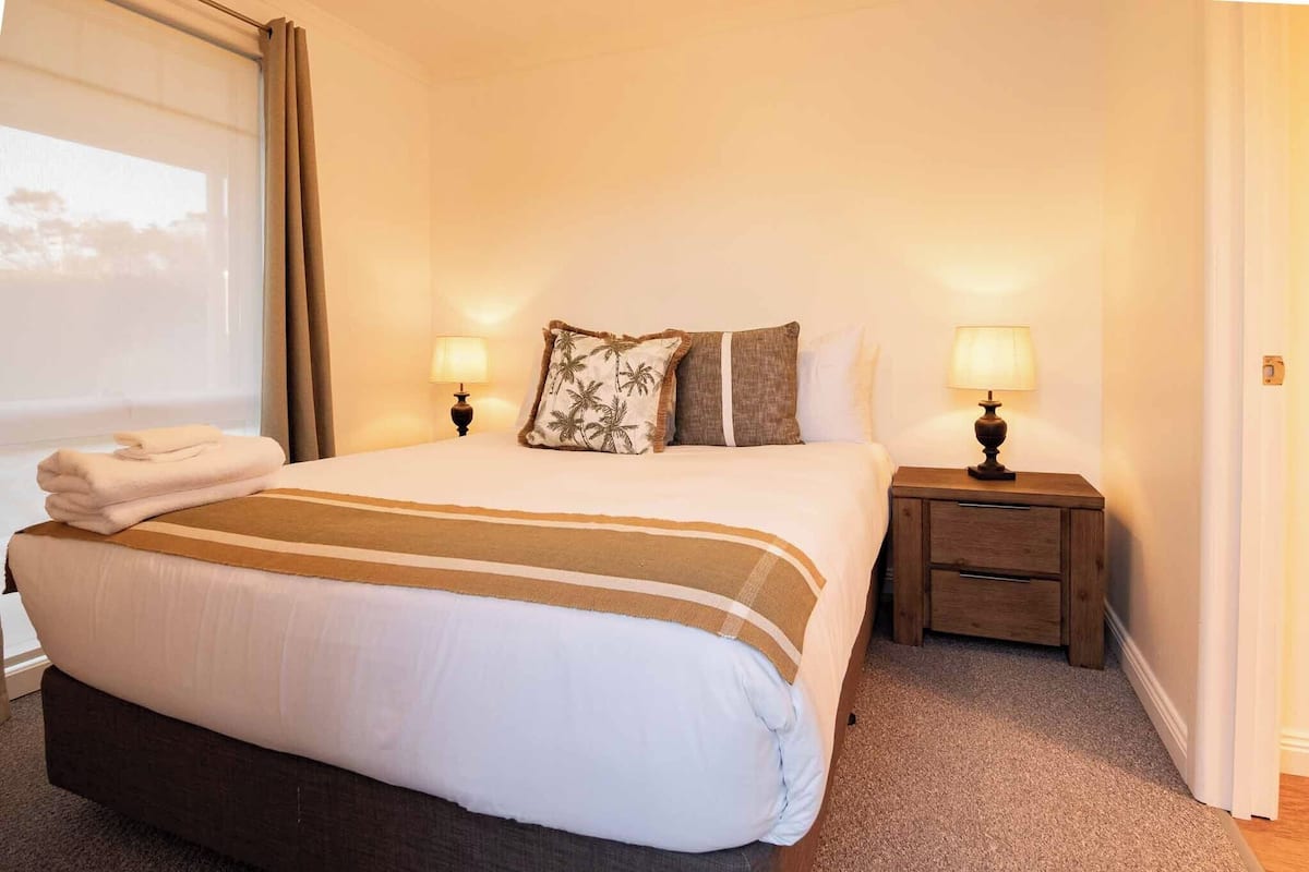 Quarto de hotel com uma cama grande, duas mesas de cabeceira com luminárias, uma janela com cortinas e vista para o exterior.