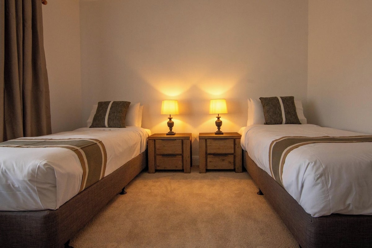 Quarto de hotel com duas camas, cada uma com mesa de cabeceira e luminária.