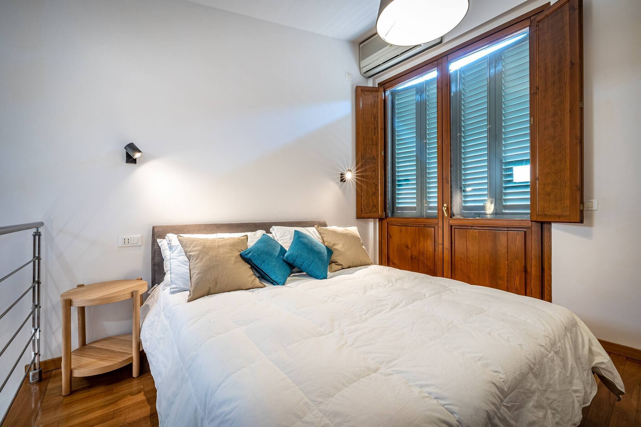 Quarto com cama, guarda-roupa de madeira, mesa de cabeceira e um banquinho.