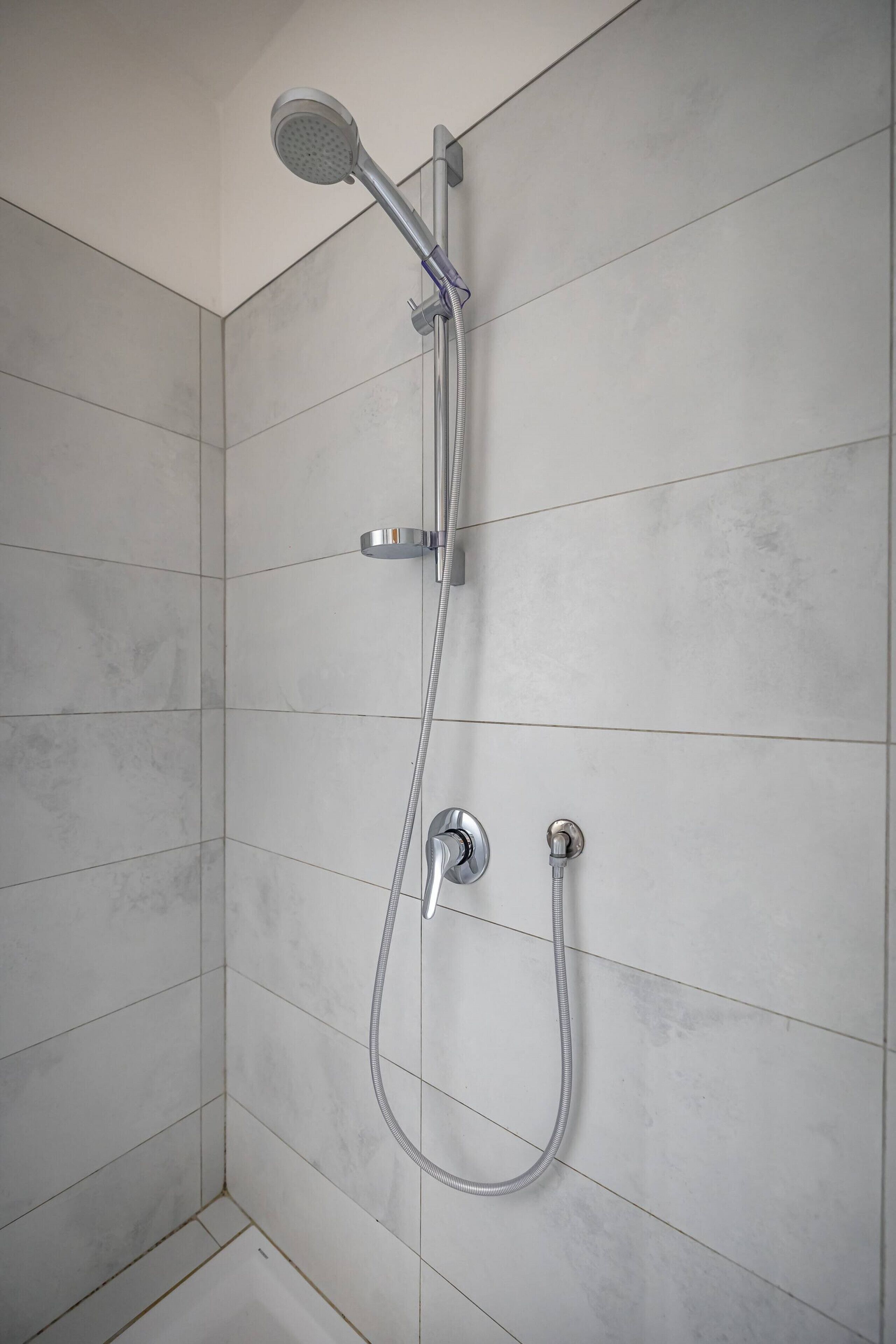 Um chuveiro com um chuveiro manual e uma torneira fixa num banheiro revestido com azulejos.