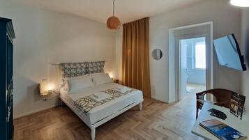 Quarto com cama, mesa de cabeceira, abajur, cortinas e porta que dá para outro cômodo.