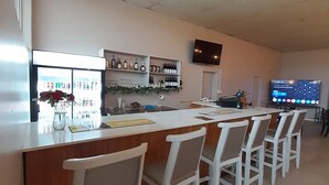 Uma área de bar com um balcão comprido, várias prateleiras com garrafas, uma televisão e algumas cadeiras.