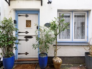 Um edifício branco com porta e janela azuis, plantas em vasos na frente, e uma placa com a inscrição 'DOLLS & COTTAGE'.