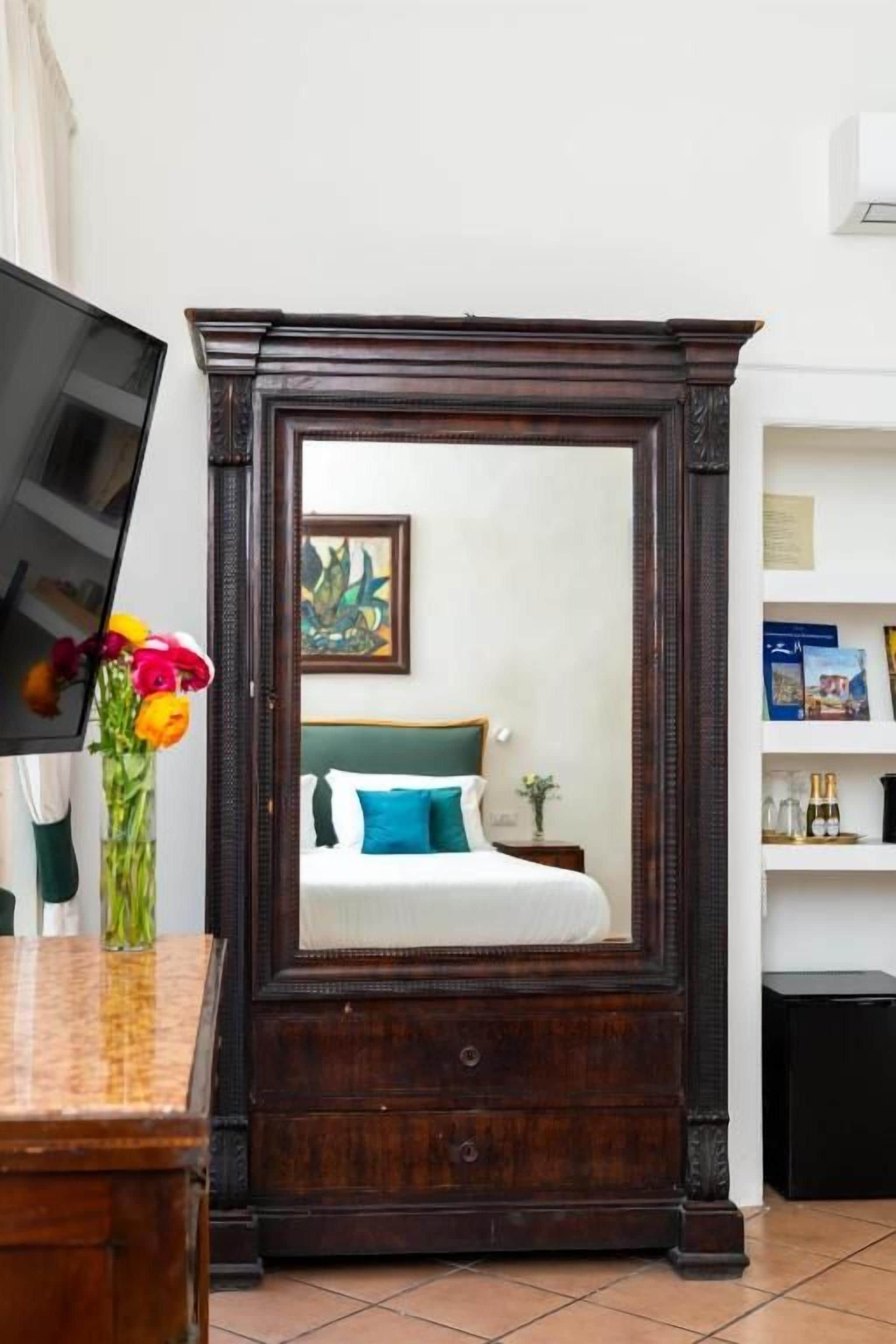 Quarto com espelho grande, cama com roupa de cama azul, mesa de cabeceira e um vaso com flores.