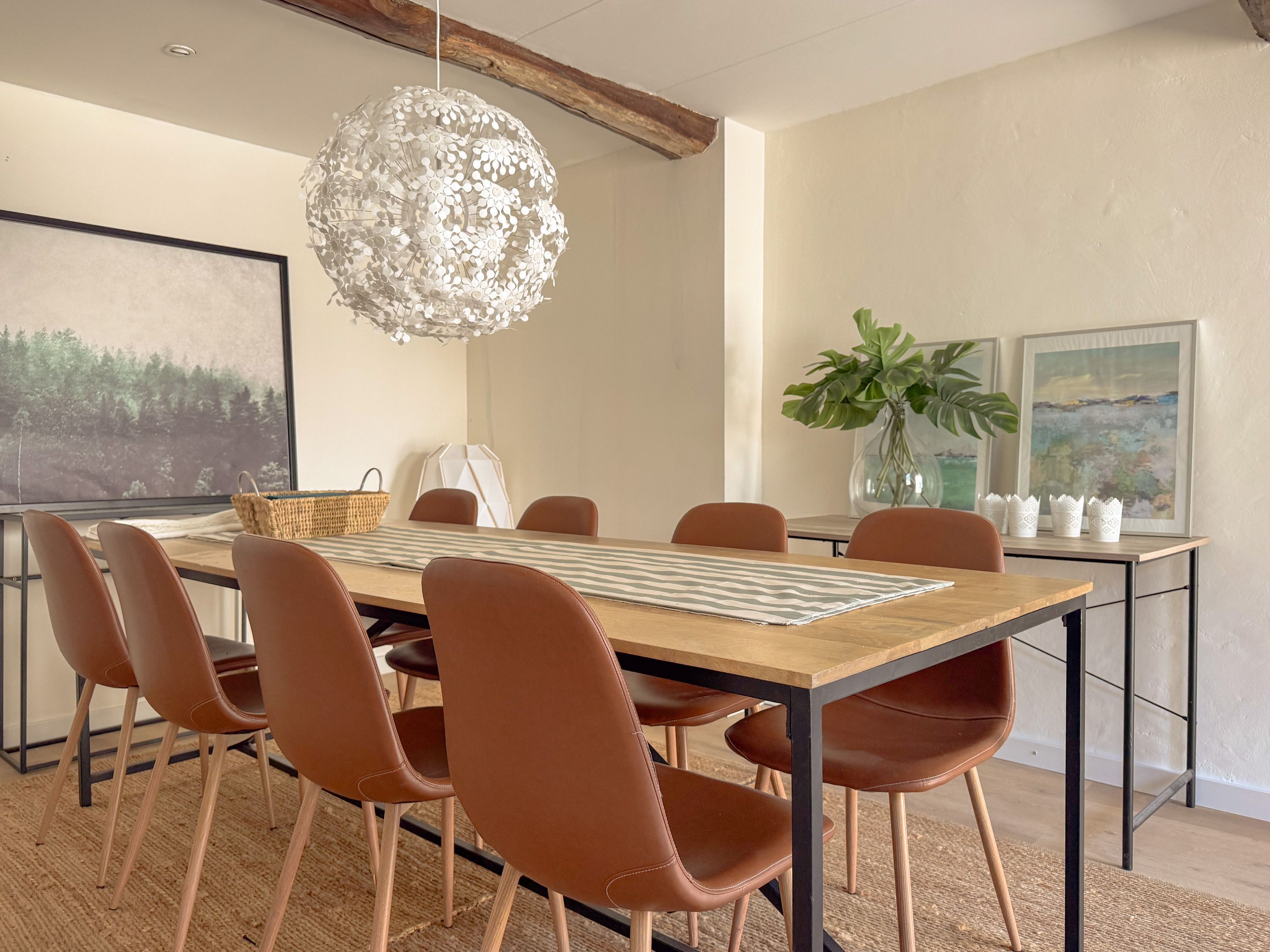 Uma área de jantar com uma mesa de madeira, cadeiras marrons, luminária pendente e quadros nas paredes.
