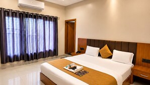 Quarto de hotel com uma cama grande, um ventilador de teto, ar condicionado, mesa de cabeceira de madeira e porta de banheiro.