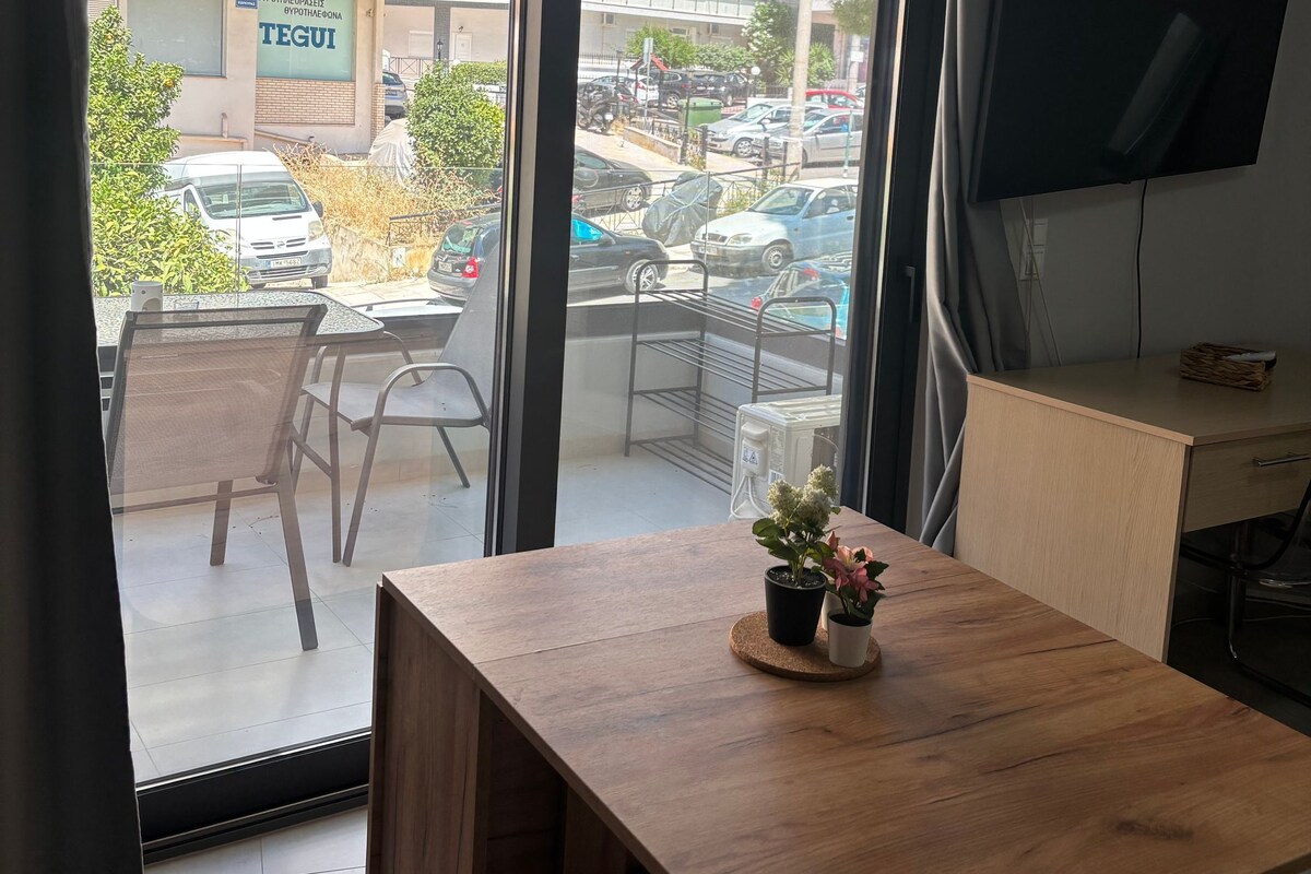 Quarto com uma mesa de madeira, um vaso de planta, uma TV e vista para a rua com carros estacionados e um edifício com uma placa.