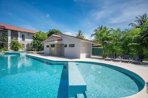 Piscina com um pequeno edifício ao lado, cercada por palmeiras e um céu azul límpido.