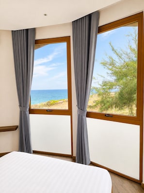 Quarto com uma grande janela, cortinas, cama e vista para a praia.