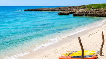 Uma praia com águas turquesa cristalinas e um afloramento rochoso ao longe. Várias pranchas de surf coloridas estão dispostas na areia.