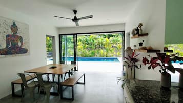 Uma área de refeições moderna com uma mesa, cadeiras, ventilador de teto e vista para uma piscina e vegetação lá fora.