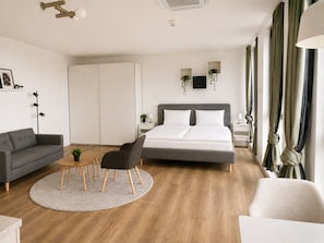 Quarto de hotel moderno com cama, sofá, pequena mesa com cadeiras, guarda-roupa e janela com cortinas.