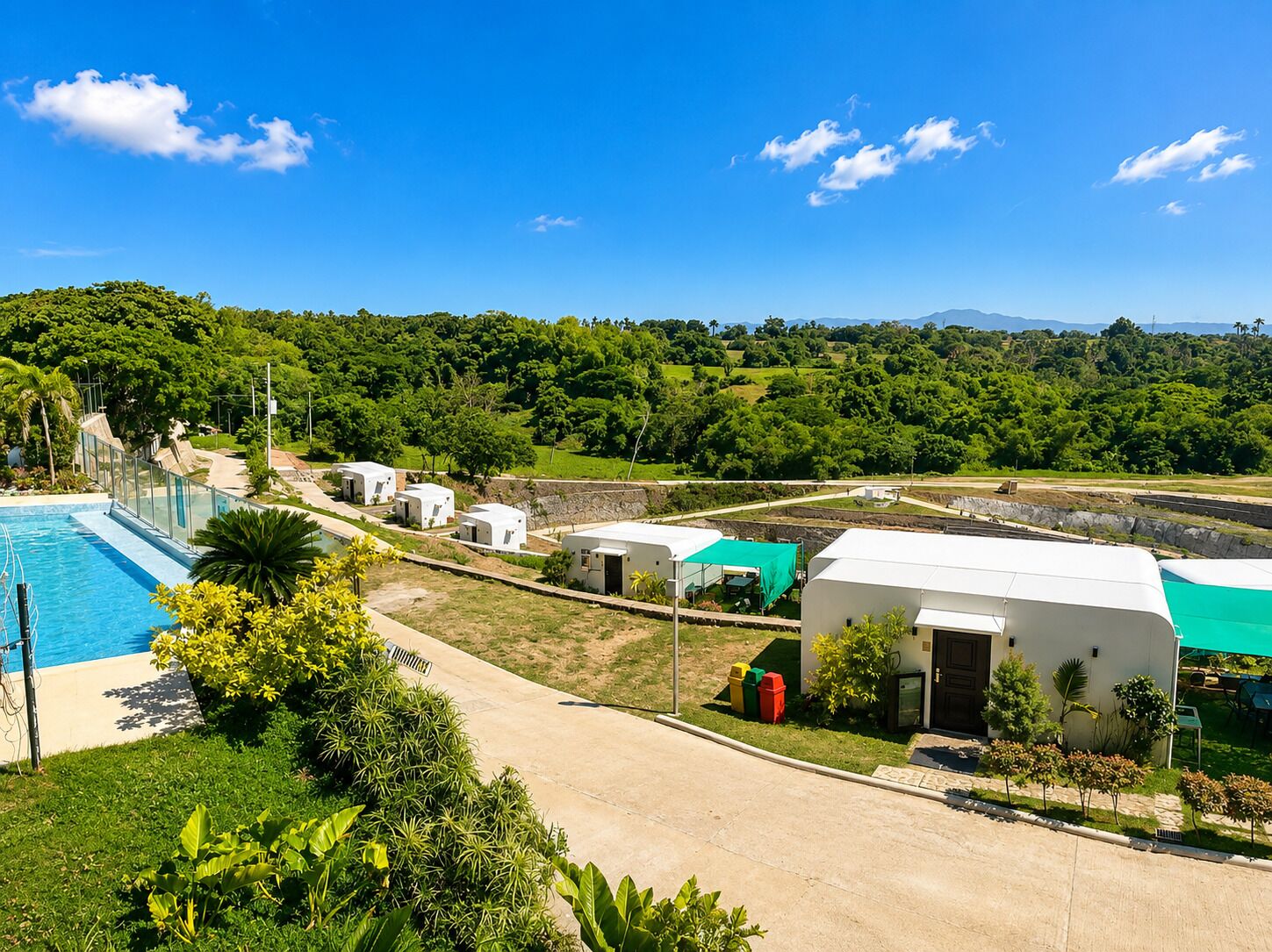 Área de piscina com céu claro, rodeada por vegetação e edifícios brancos com telhados verdes.