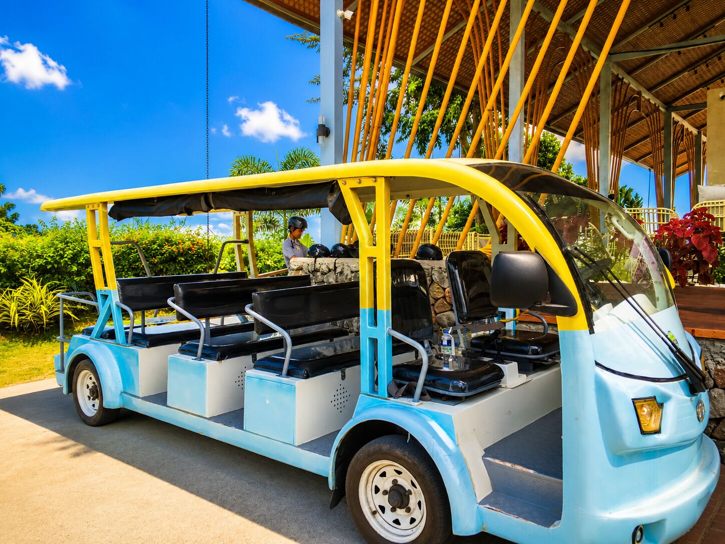 Um veículo de três rodas azul e amarelo com área de estar coberta, estacionado sob uma estrutura com telhado de bambu.