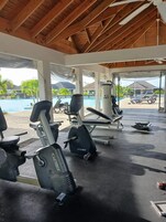 Uma área de ginástica coberta ao ar livre com equipamentos de exercício, piscina e espreguiçadeiras.