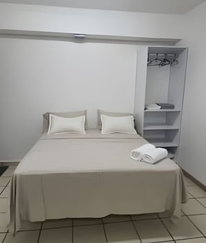 Quarto individual com uma colcha branca, duas almofadas, uma toalha dobrada e um guarda-roupa com espelho.