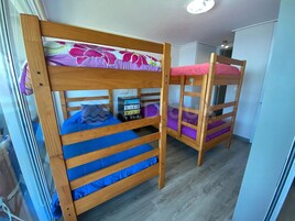 Quarto com duas beliches, uma pequena mesa de cabeceira com abajur e uma janela.