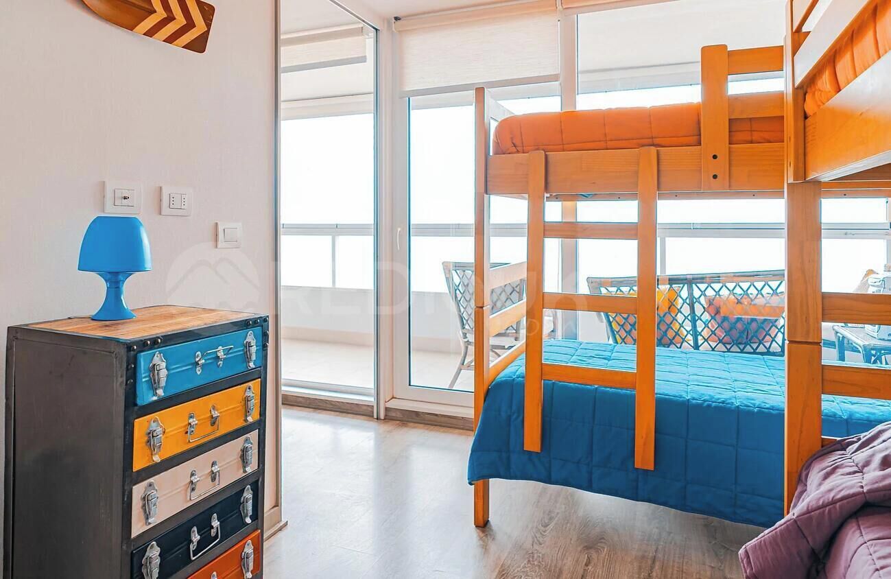 Uma cama beliche com colcha azul, uma cômoda com várias gavetas e uma janela com vista para o exterior.