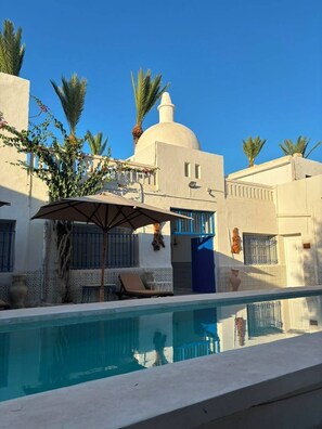 Uma área de piscina com espreguiçadeira e guarda-sol, cercada por edifícios com paredes brancas e detalhes azuis.