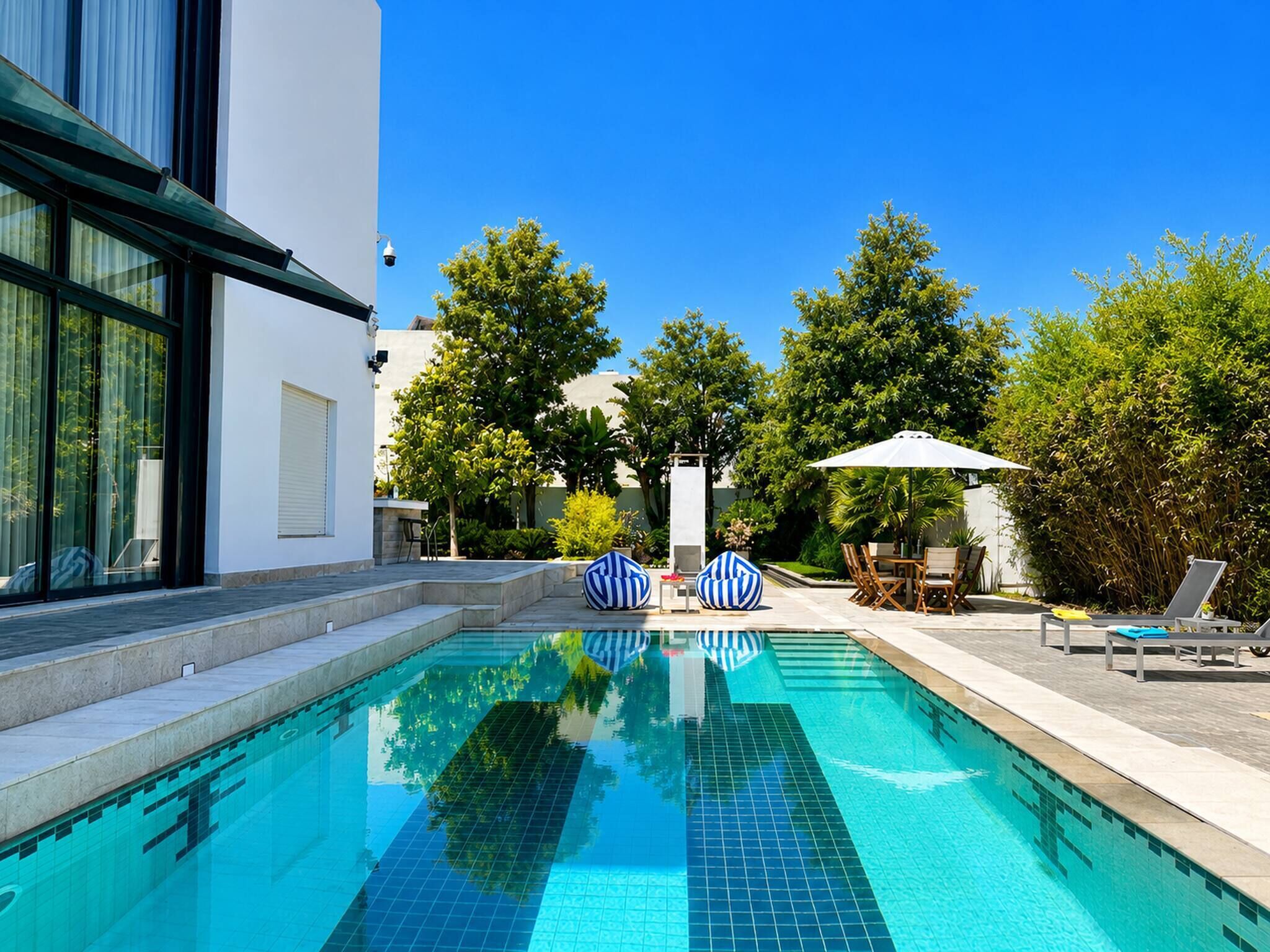 Piscina com espreguiçadeiras e guarda-sóis, cercada por árvores e um edifício moderno.