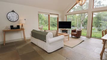 Uma sala de estar com um sofá branco, televisão, mesa de centro de madeira e uma janela grande que oferece vista para o verde.