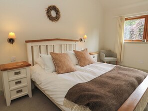 Quarto com cabeceira de madeira, cama branca, duas almofadas, manta marrom, mesa de cabeceira com gavetas, uma cadeira, janela com cortinas e luminária na parede.