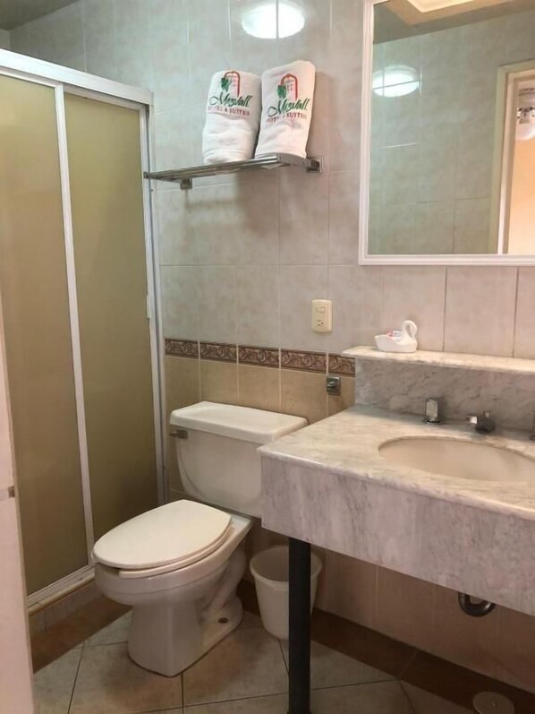 Casa de banho com sanita, lavatório e chuveiro. Há toalhas numa prateleira acima do lavatório.
