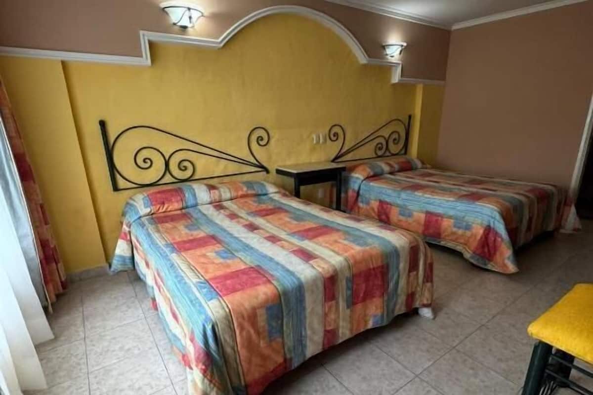 Quarto com duas camas, uma mesinha e luminária fixada na parede.