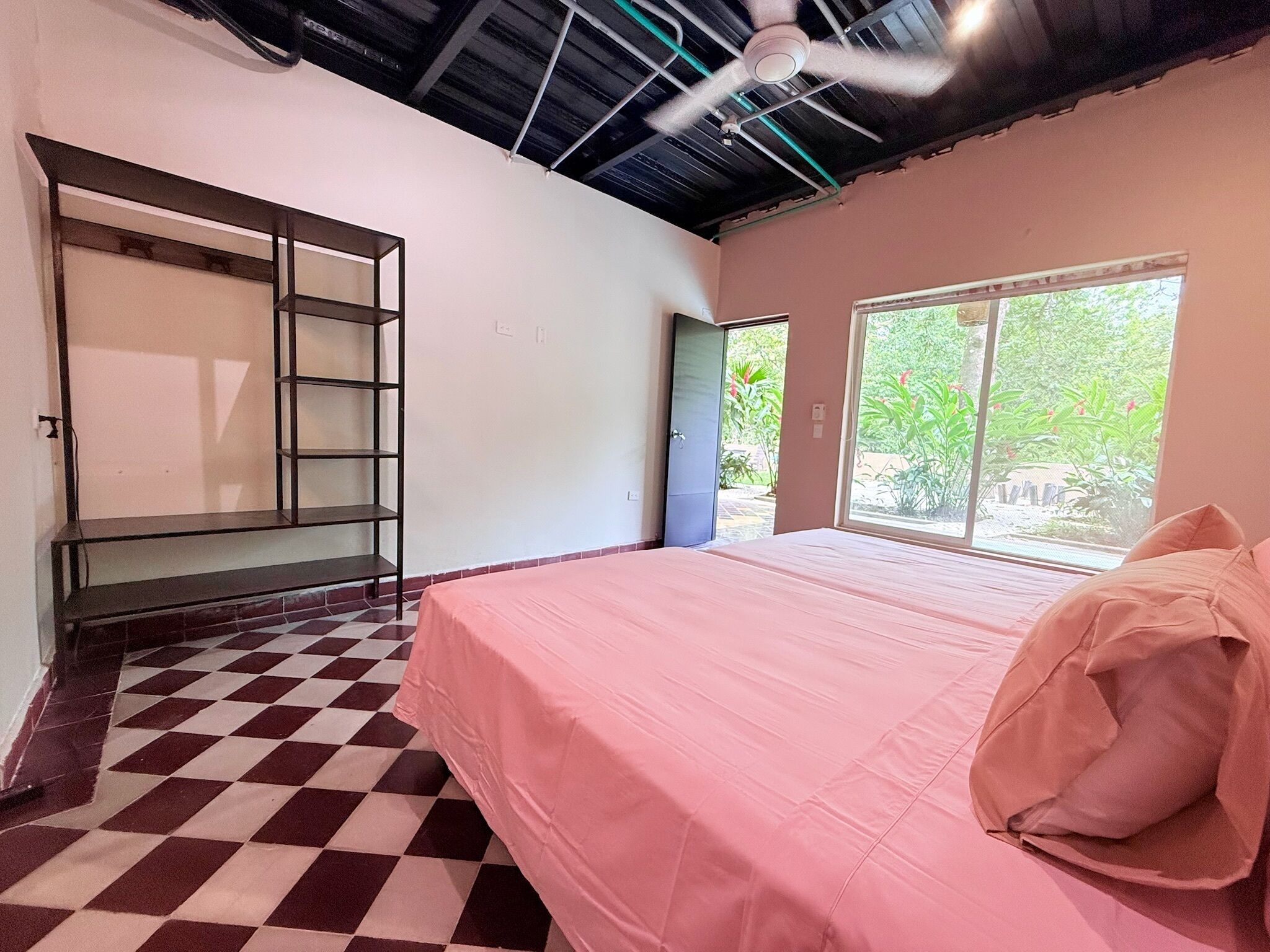Quarto com piso xadrez, cama com colcha rosa, estante de metal e janela com vista para vegetação.