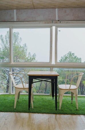 Quarto com mesa, duas cadeiras, uma janela grande e vista para árvores.