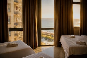 Quarto de hotel com duas camas, uma janela grande com cortinas e vista para a paisagem urbana e para a água.