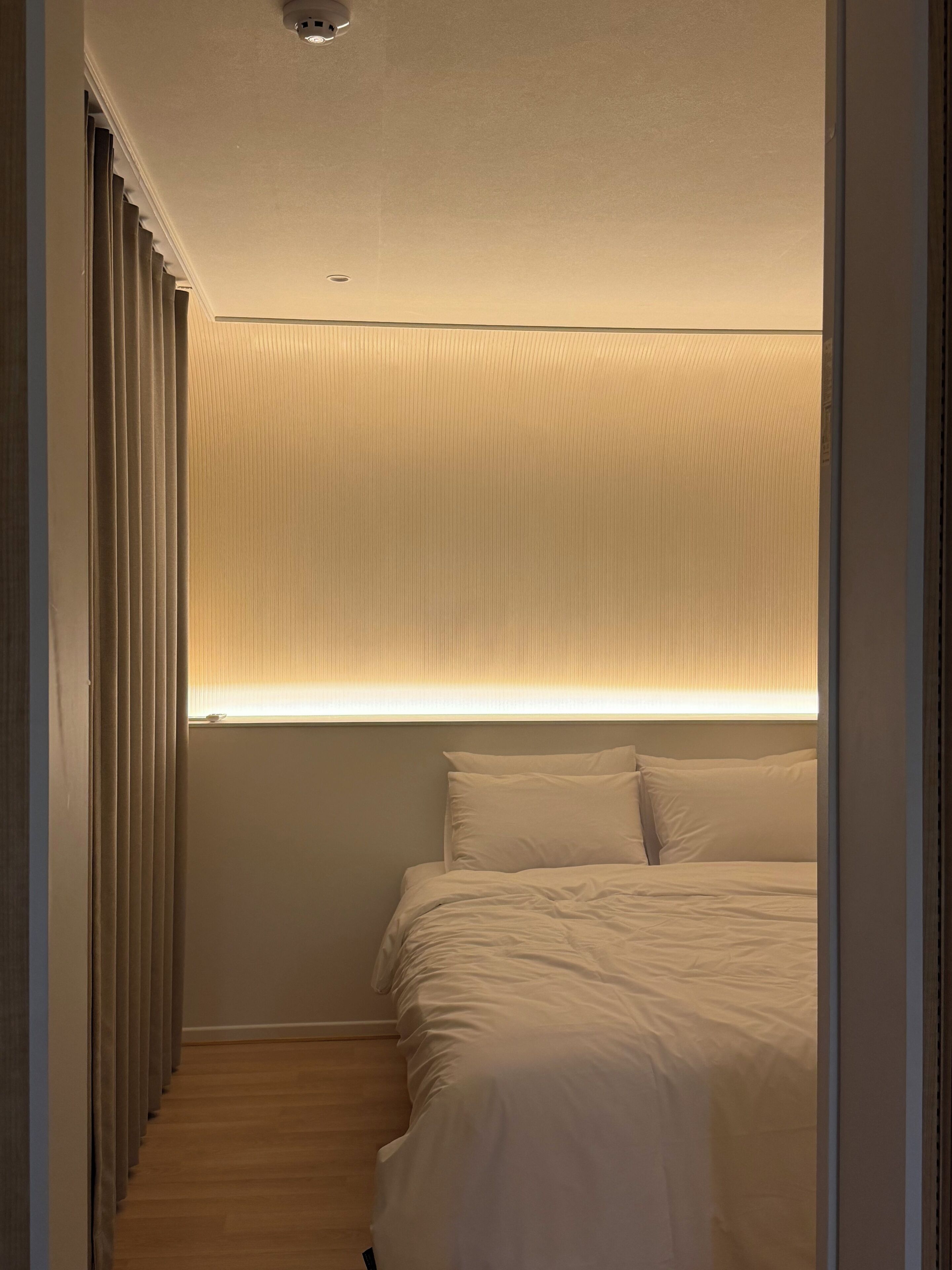Quarto com cama, janela com persianas, luminária no teto e piso de madeira.
