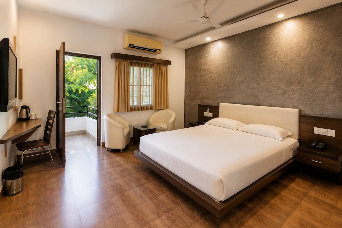 Quarto de hotel com uma cama grande, uma escrivaninha com cadeira, uma mesinha com duas cadeiras, ar condicionado e vista para o verde pela janela.