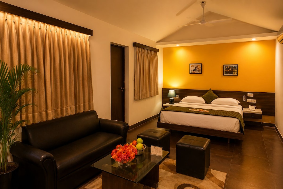 Quarto de hotel com uma cama, um sofá, uma mesa de centro com flores e uma mesinha de cabeceira com abajur.