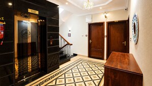 Um lobby de hotel com um elevador preto, portas de madeira, um candelabro e um piso com padrão.