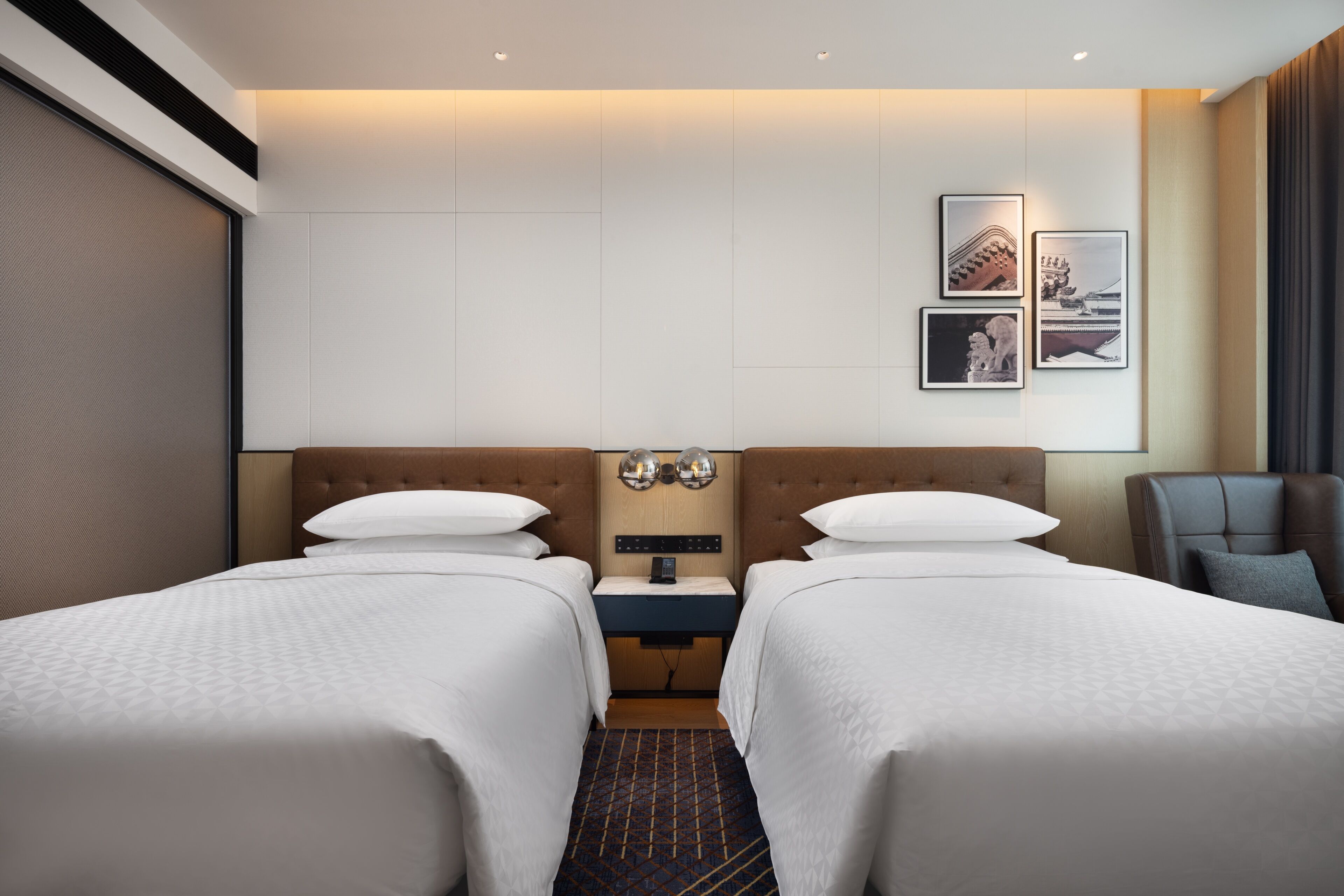 Quarto de hotel com duas camas, uma mesa de cabeceira, luminárias fixadas na parede e quadros emoldurados.