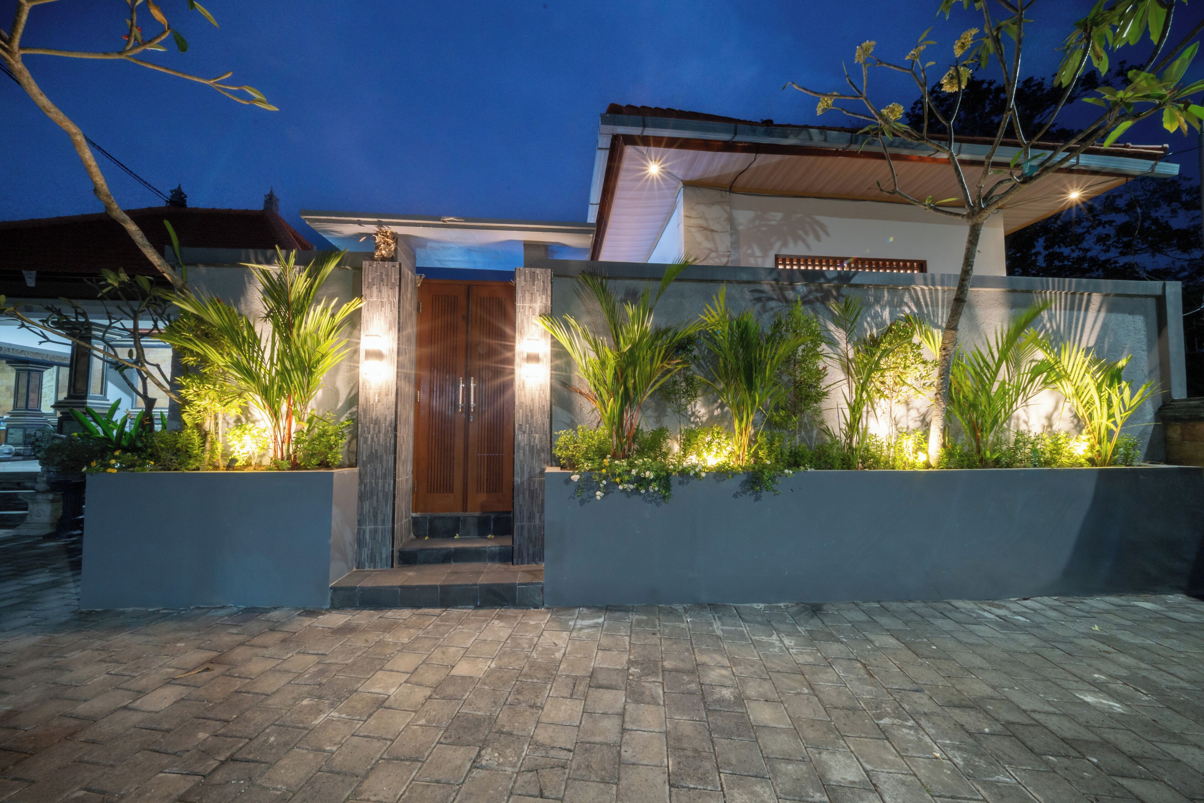 Uma casa moderna com entrada pavimentada, porta de madeira e plantas iluminadas.