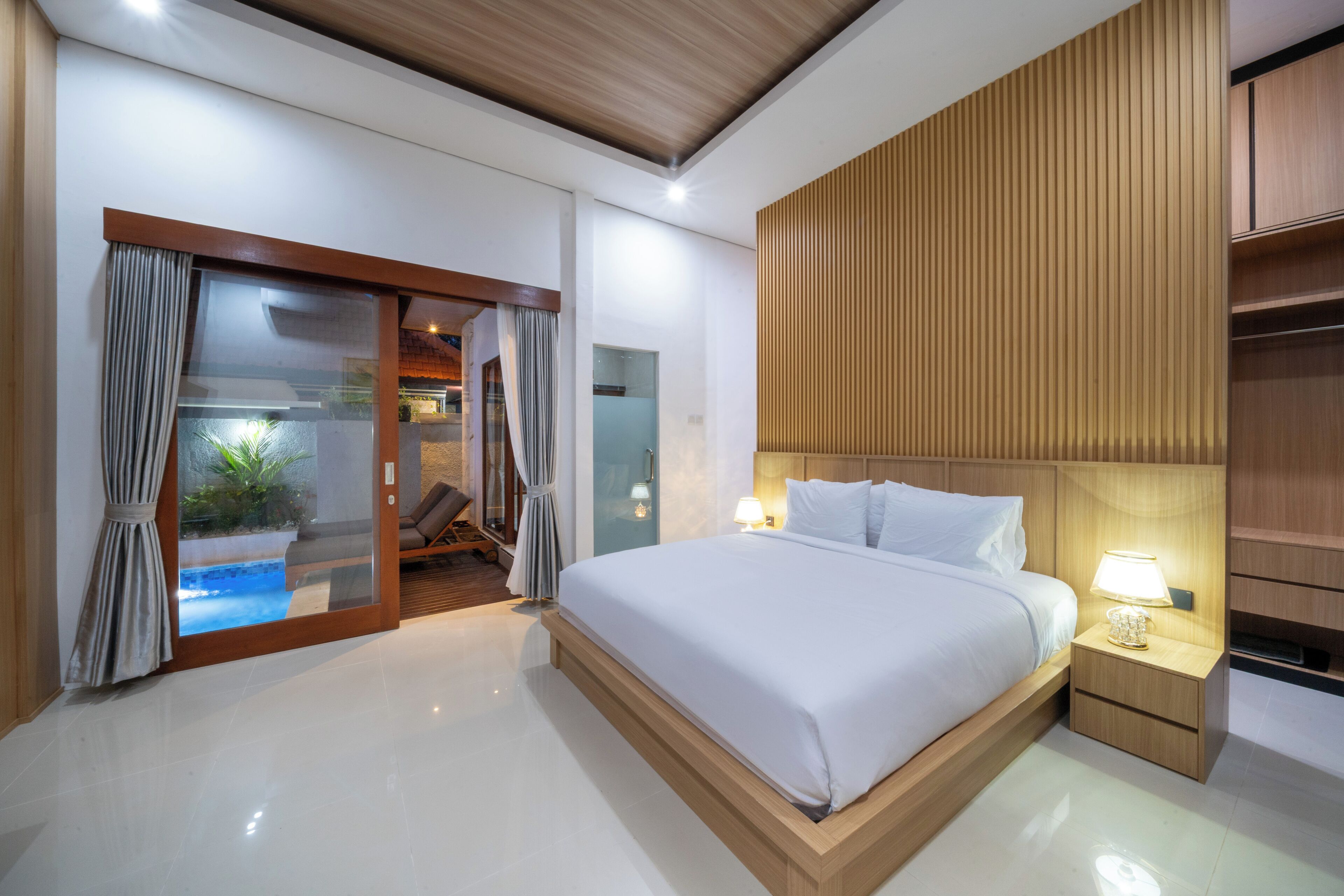 Quarto de hotel moderno com uma cama grande, mesa de cabeceira com abajur, porta de vidro deslizante para uma varanda e cabeceira de madeira.