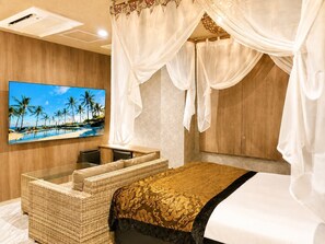 Quarto de hotel com cama, televisão exibindo uma cena de praia, guarda-roupa de madeira e uma cadeira de vime.