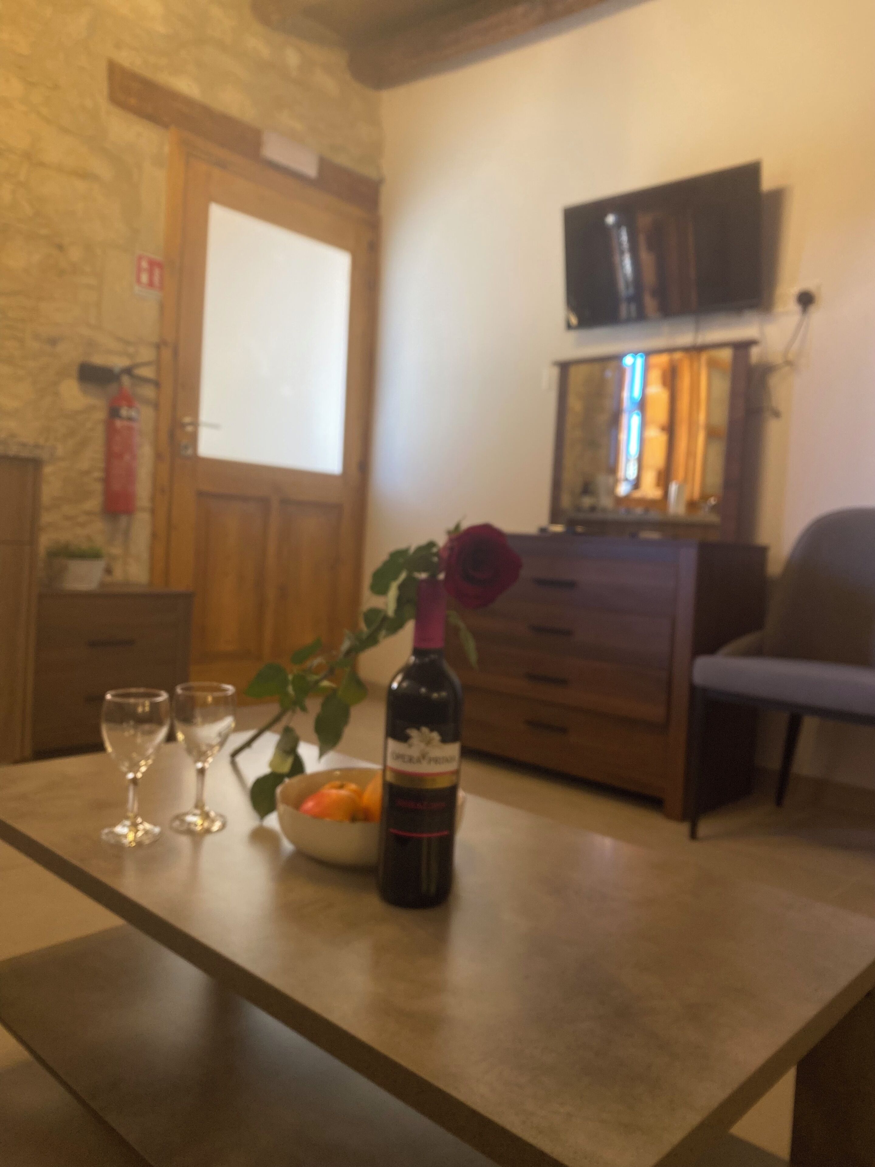 Quarto com parede de pedra, porta de madeira, televisão na parede, mesa com garrafa de vinho, copos e uma tigela de frutas.