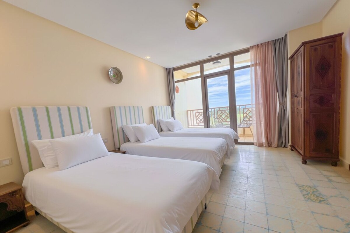Quarto de hotel com duas camas, um guarda-roupa de madeira, varanda com vista e chão ladrilhado.