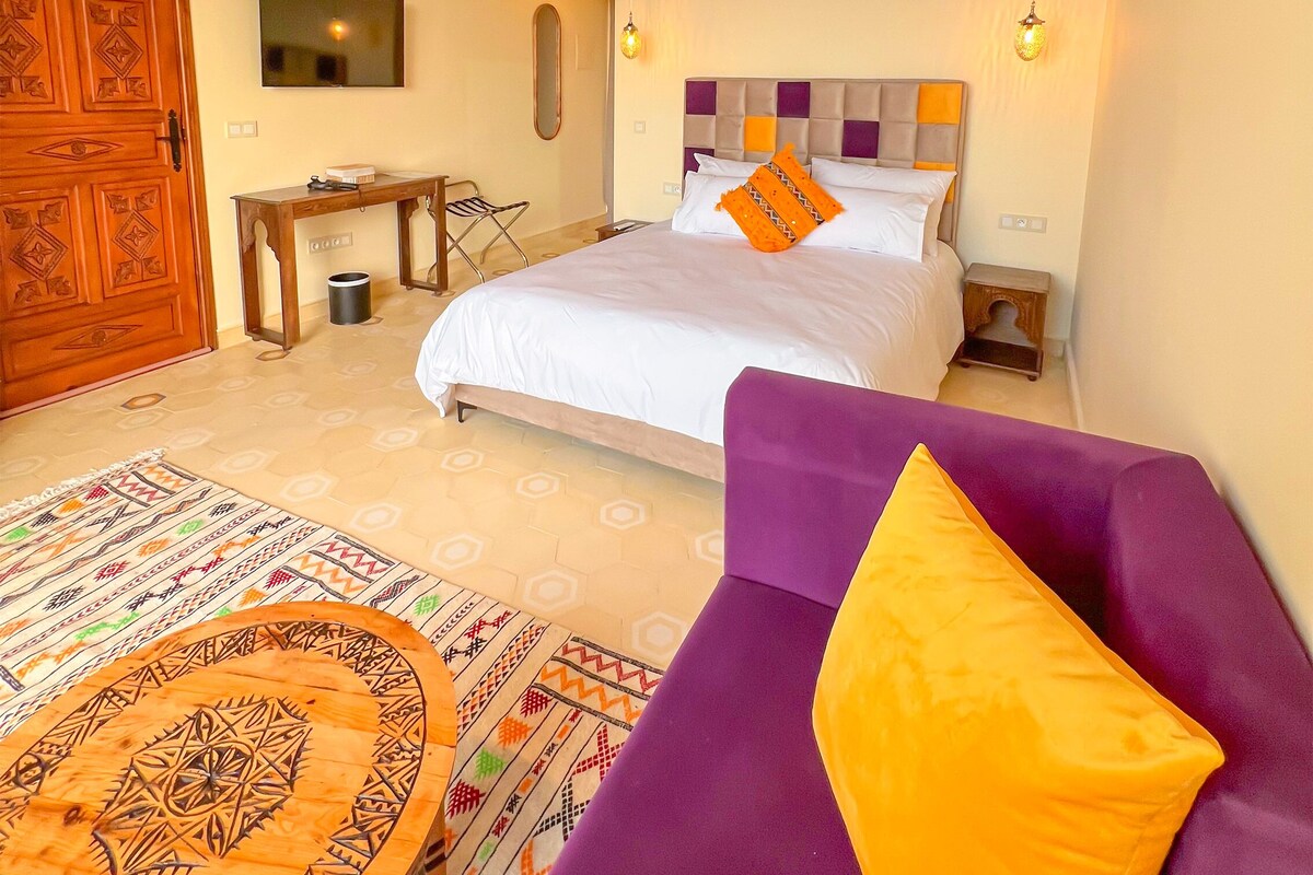 Quarto com cama, sofá roxo, mesa de madeira e televisão.