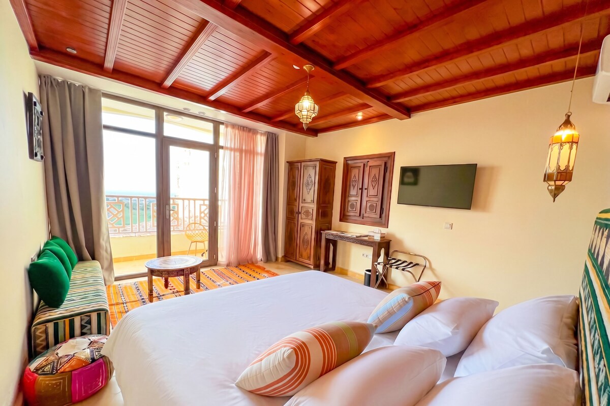 Quarto de hotel com uma cama grande, televisão, varanda com cortinas e mobiliário de madeira.